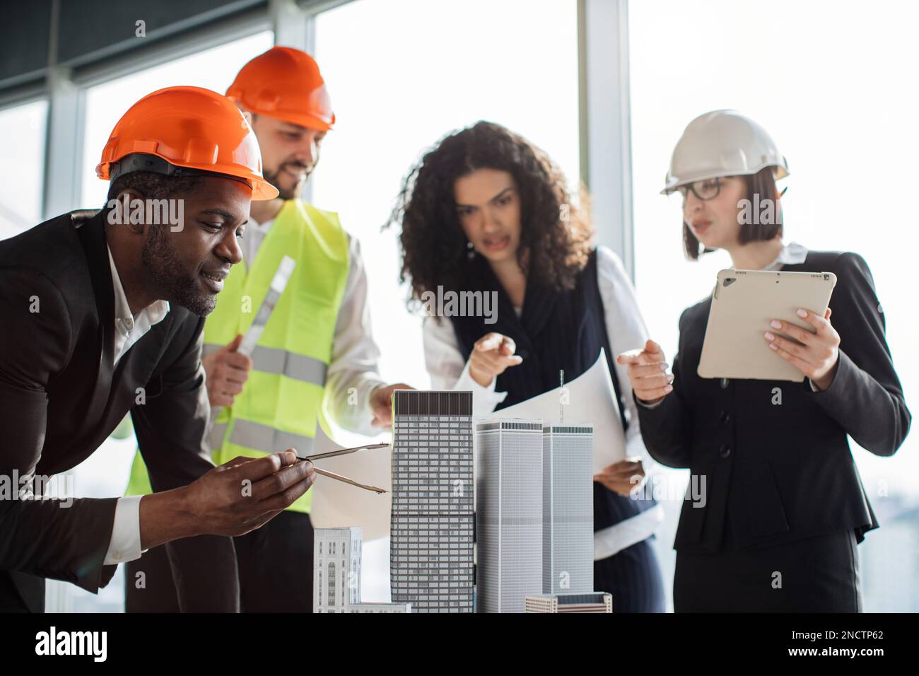 African engineer in protective helmet measuring city model using compass while colleagues ...