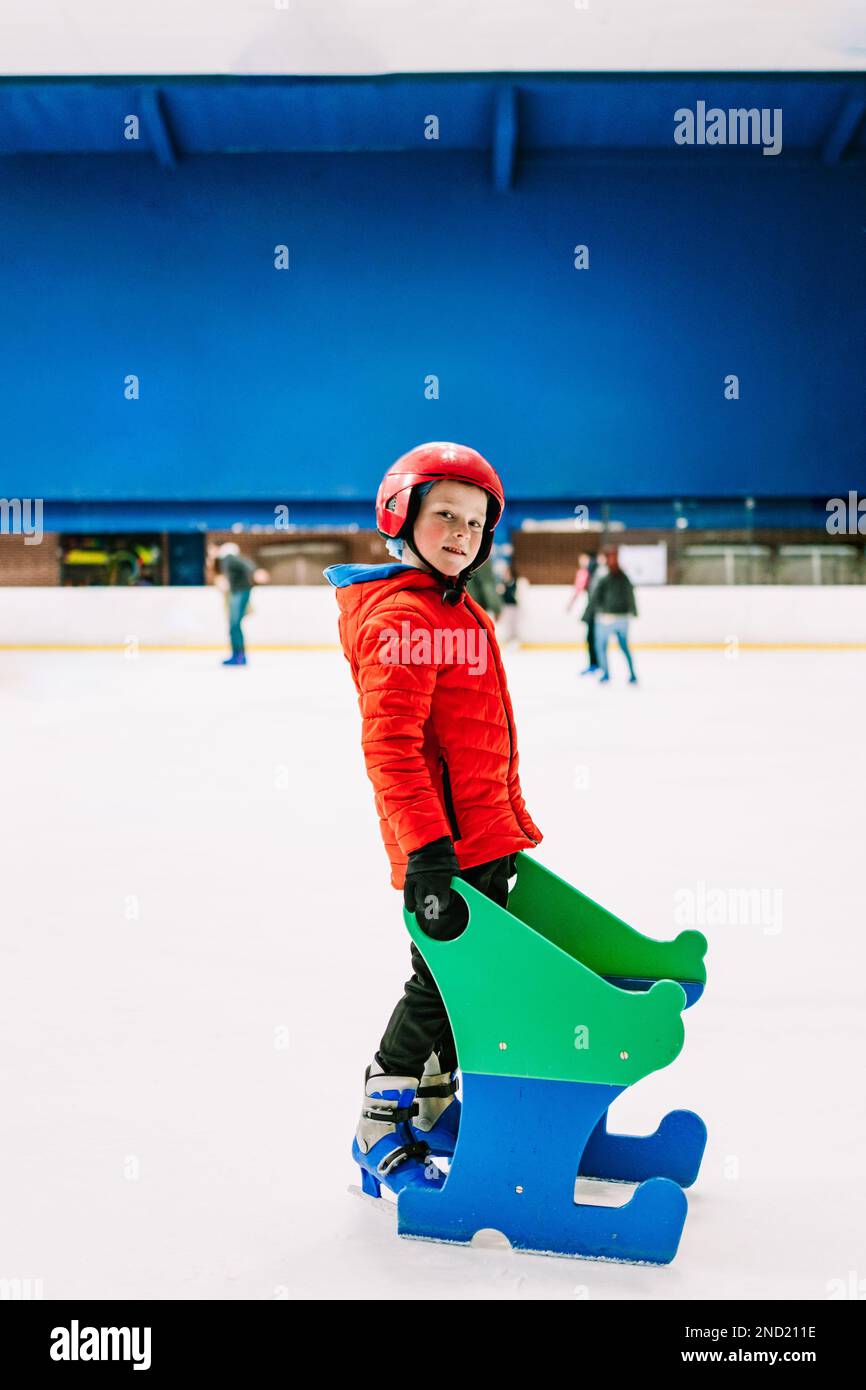 Cheerful boy in warm clothes and protective helmet learning ice skating ...