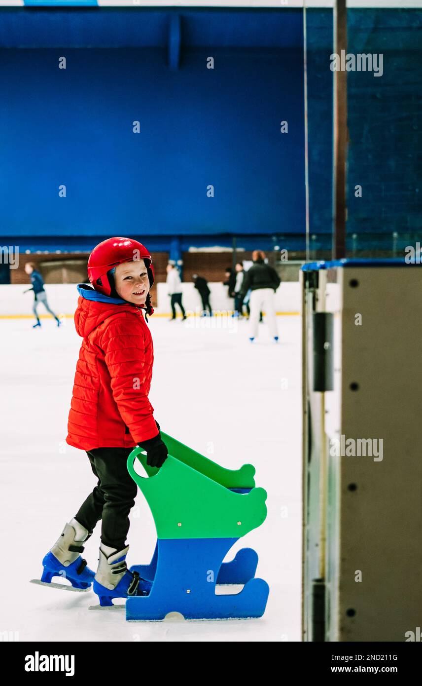 Cheerful boy in warm clothes and protective helmet learning ice skating ...