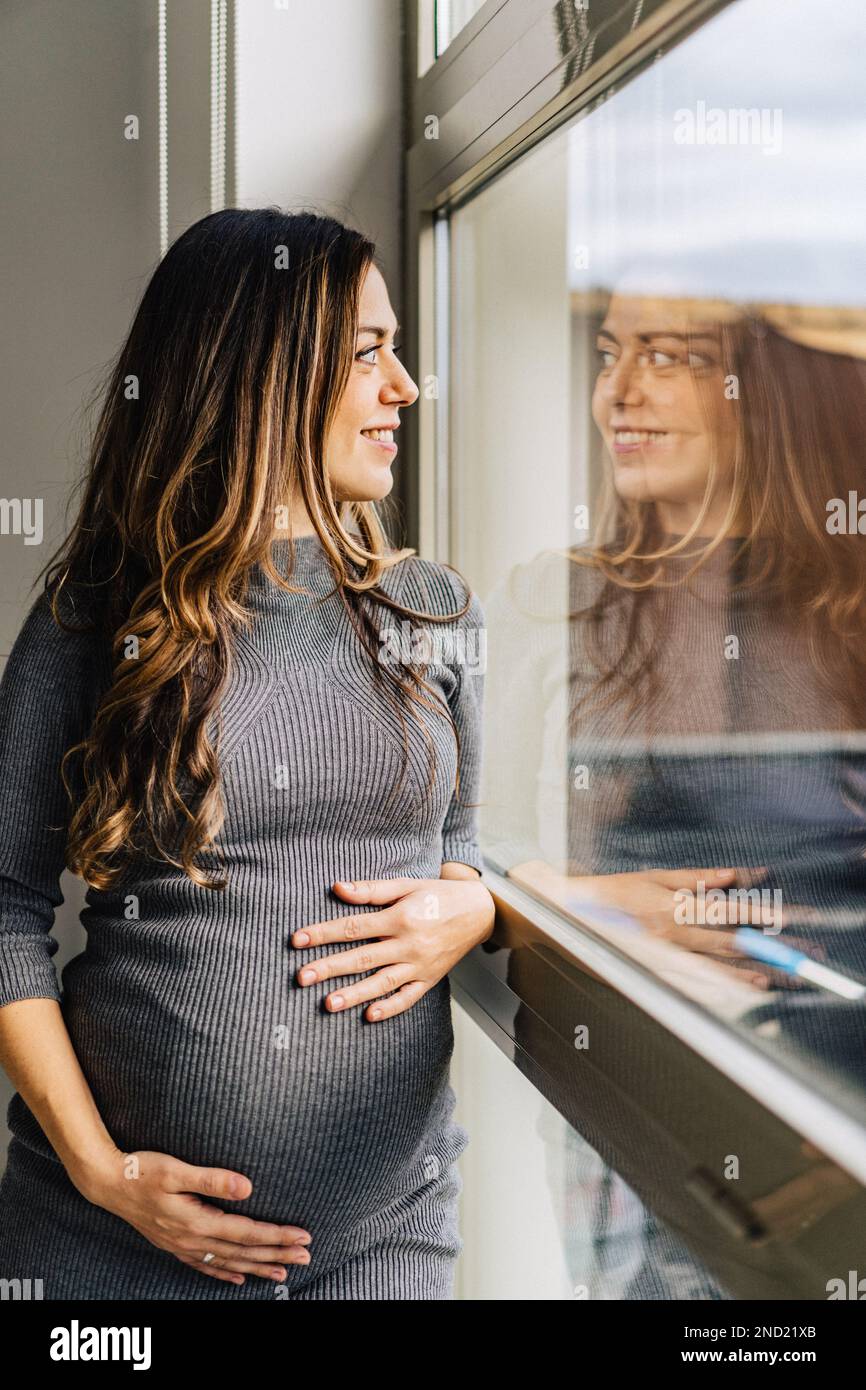 Calm female in gray dress looking out of window while touching pregnant ...