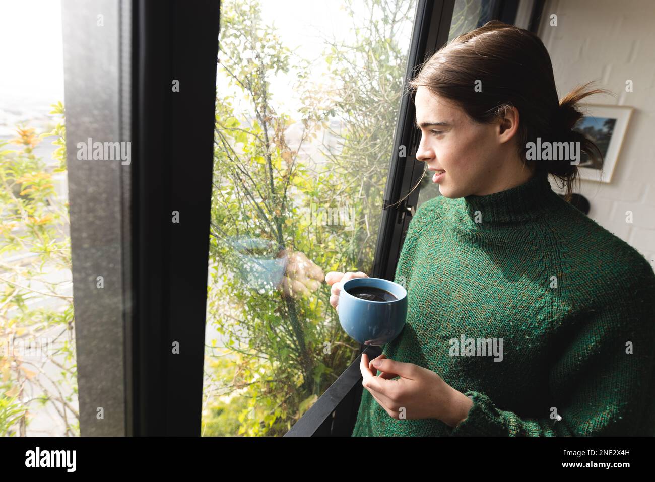 Non-binary trans woman holding coffee cup looking out of the window at ...
