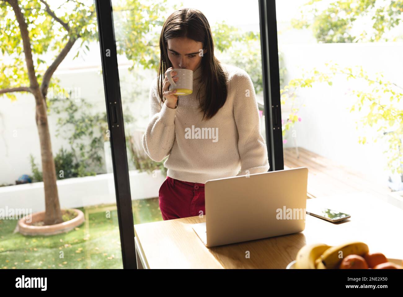 Non-binary trans woman drinking coffee and using laptop in the kitchen ...