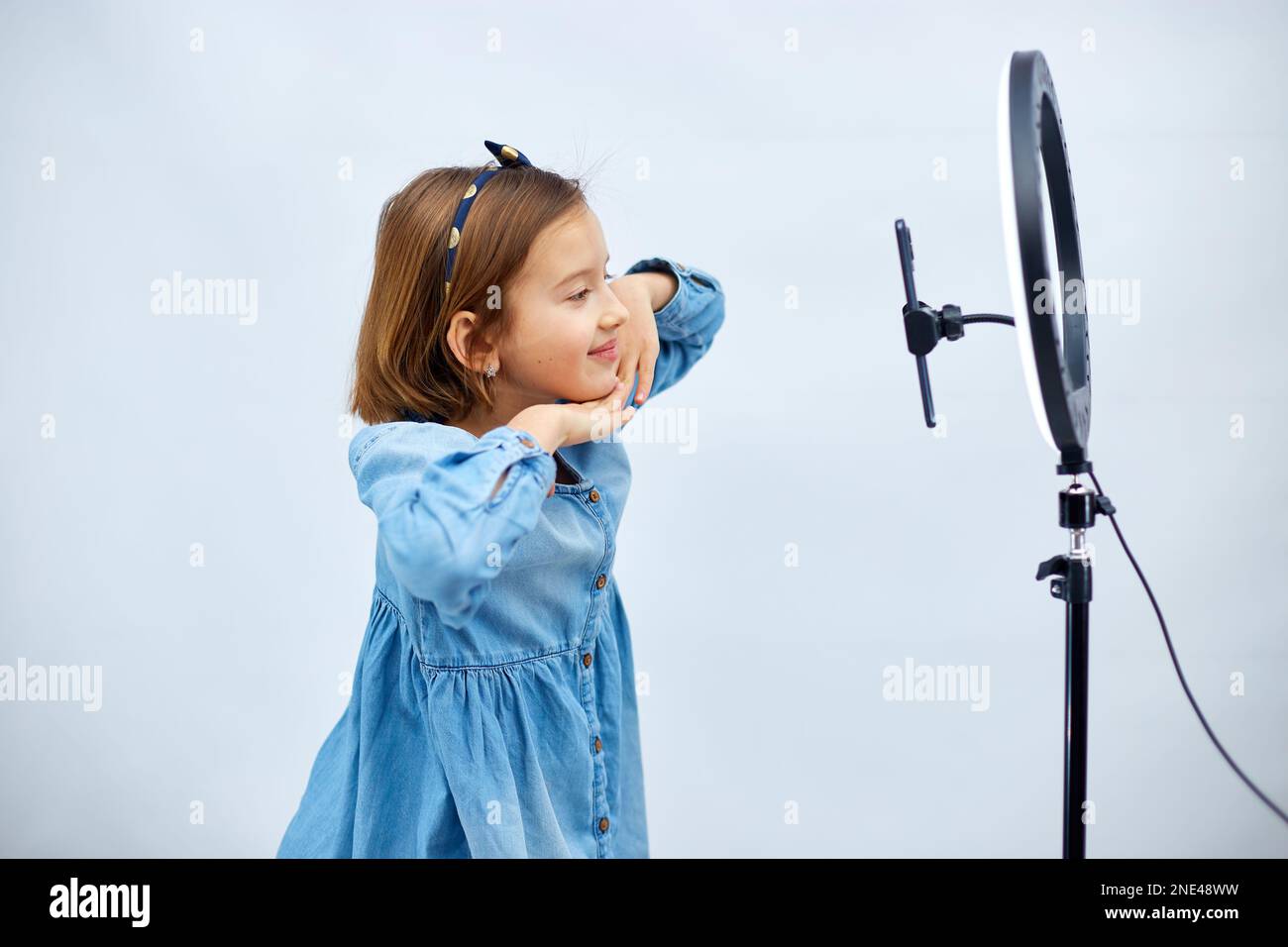 Smiling little girl blogger in casual denim dress use selfie led lamp ...
