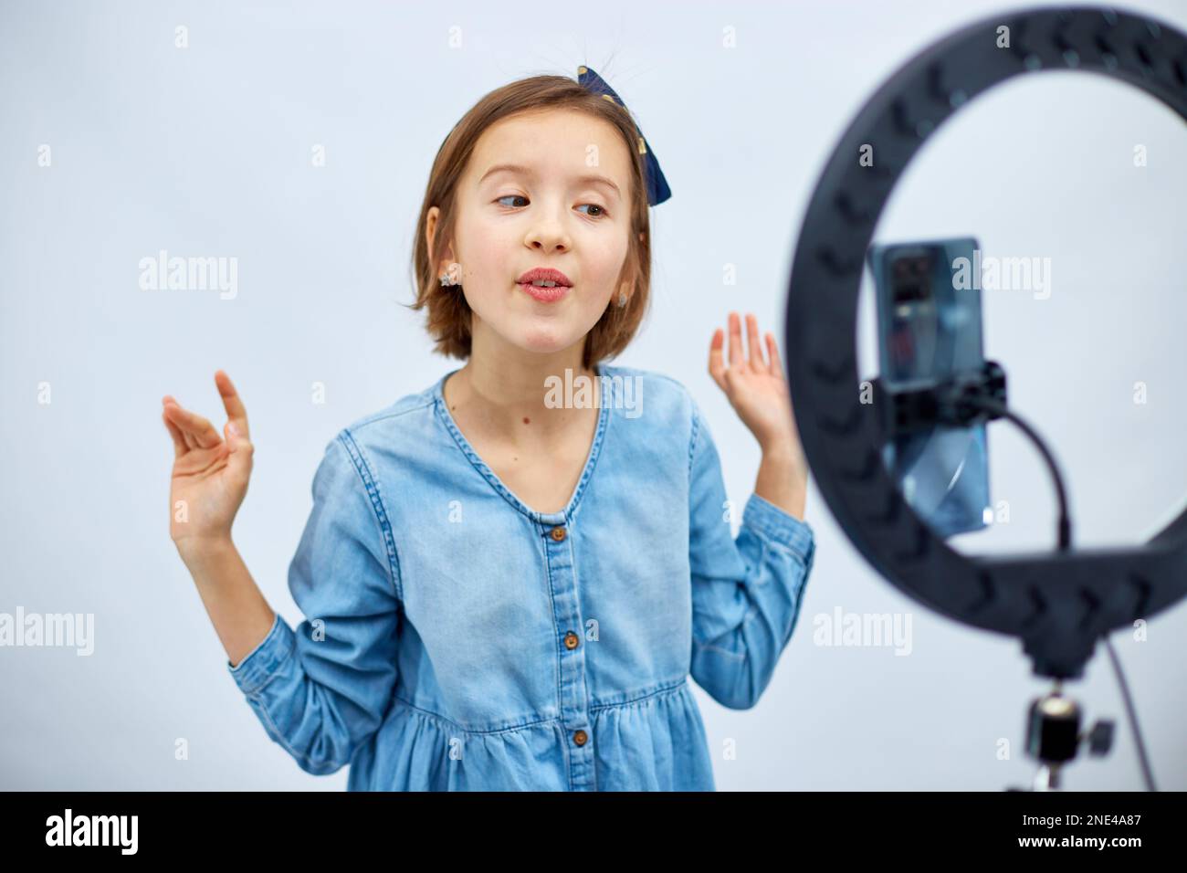 Smiling little girl blogger in casual denim dress use selfie led lamp ...