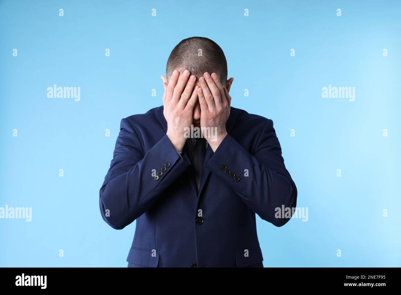 Upset man in suit closing his face with hands on light blue background ...