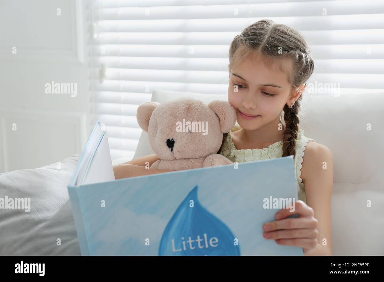 Cute little girl with teddy bear reading book on sofa at home Stock ...