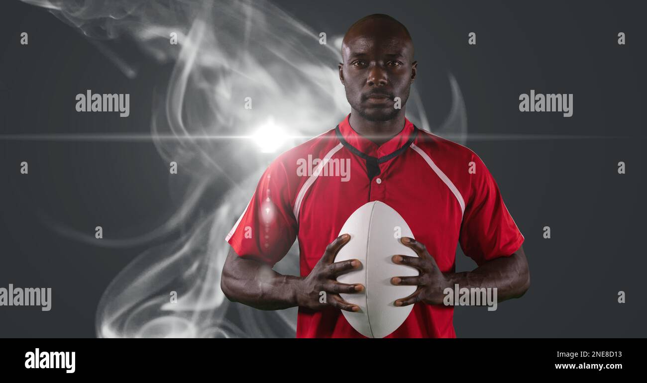African american male rugby player holding ball against smoke and light ...