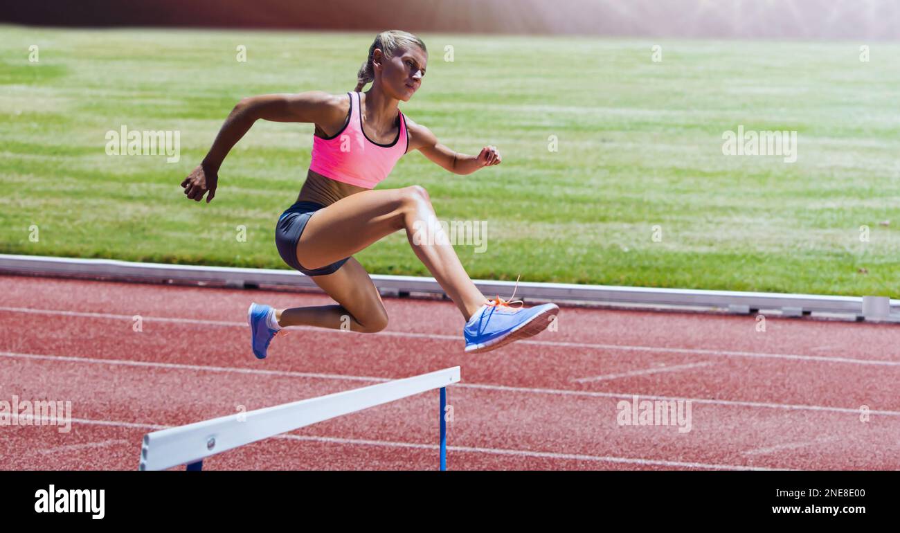 Caucasian female athlete jumping over a hurdle against sports field in ...