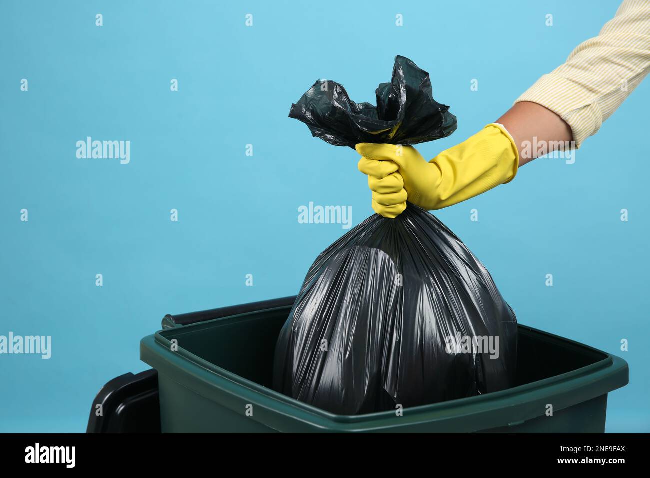 Woman throwing garbage bag into bin on light blue background, closeup ...