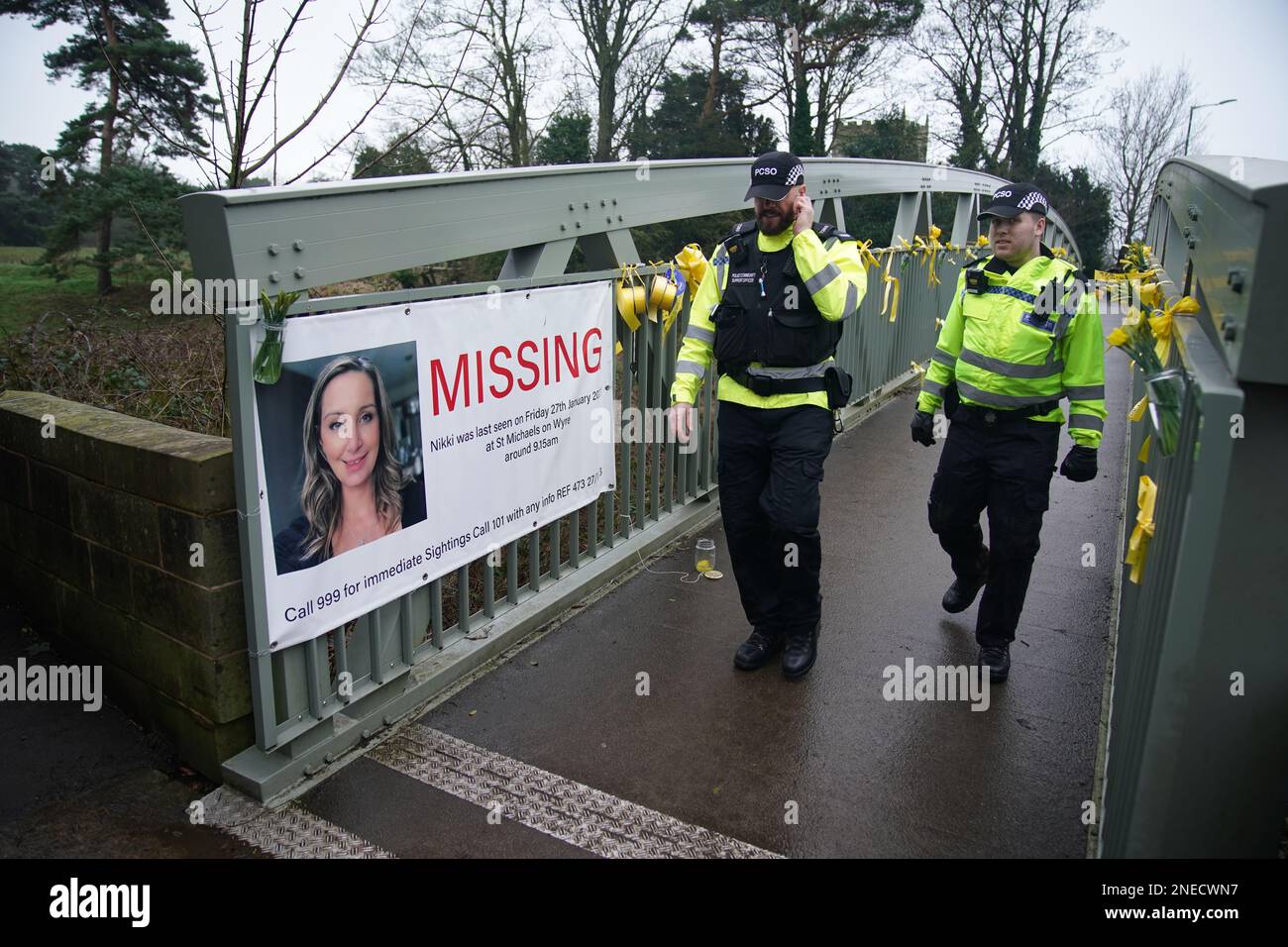 Police officers walk past a missing person appeal poster for Nicola Bulley and yellow ribbons ...