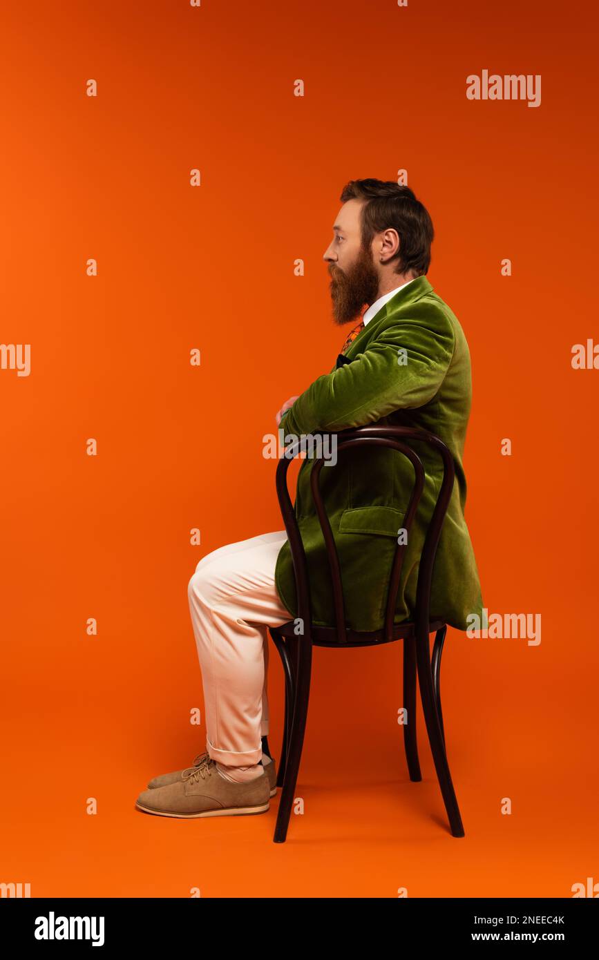 Side view of stylish man in velvet jacket sitting on wooden chair on ...