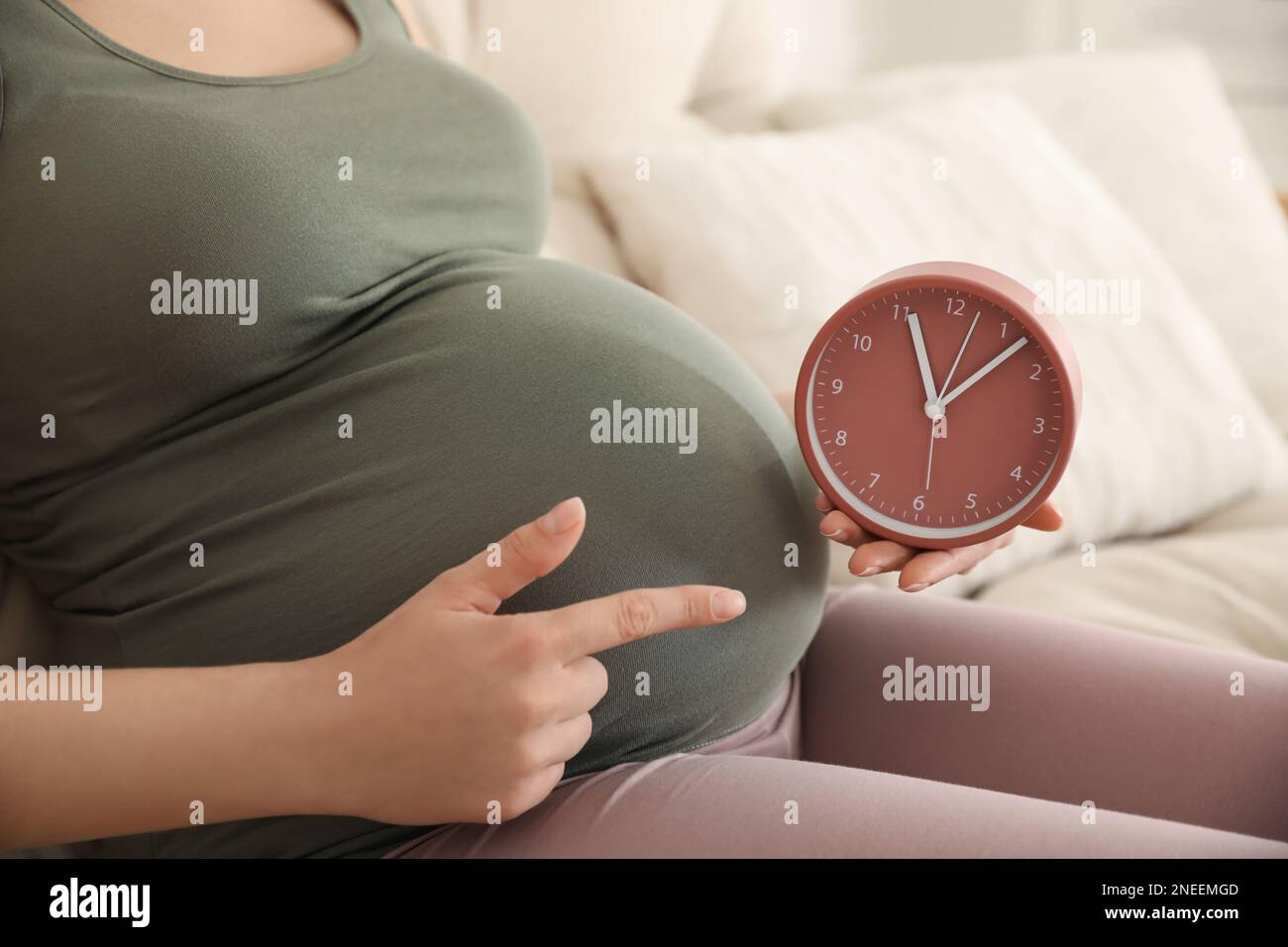 Young pregnant woman pointing at clock near her belly indoors, closeup ...