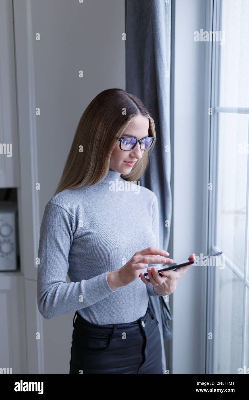 Beautiful young adult woman browsing tablet computer by the window ...