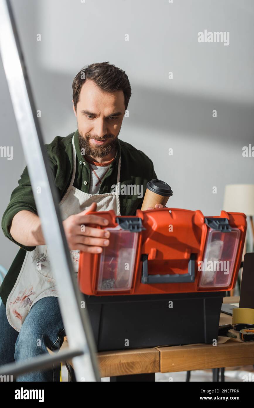 Bearded carpenter holding paper cup and opening toolbox in workshop ...