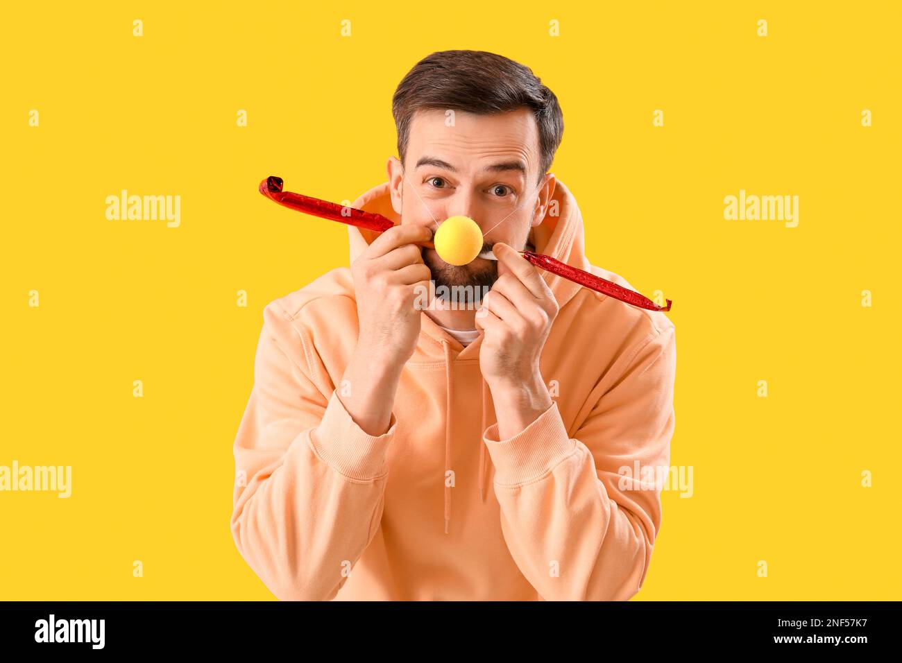 Funny young man with clown nose and party blowers on yellow background ...