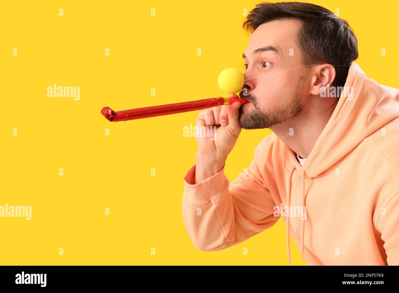 Funny young man with clown nose and party blower on yellow background ...