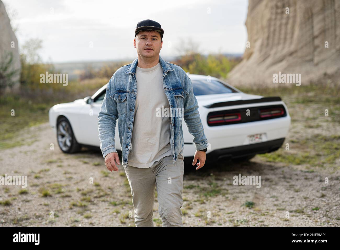 Handsome man in jeans jacket and cap walking near his white muscle car ...