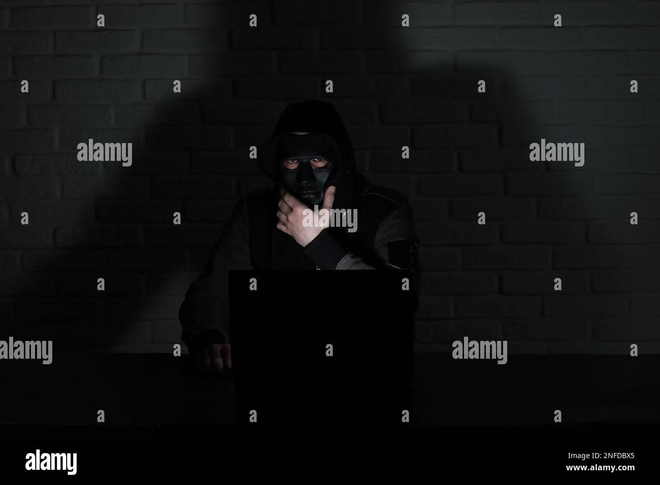 Hacker in black mask and hood at the table in front of the monitor ...