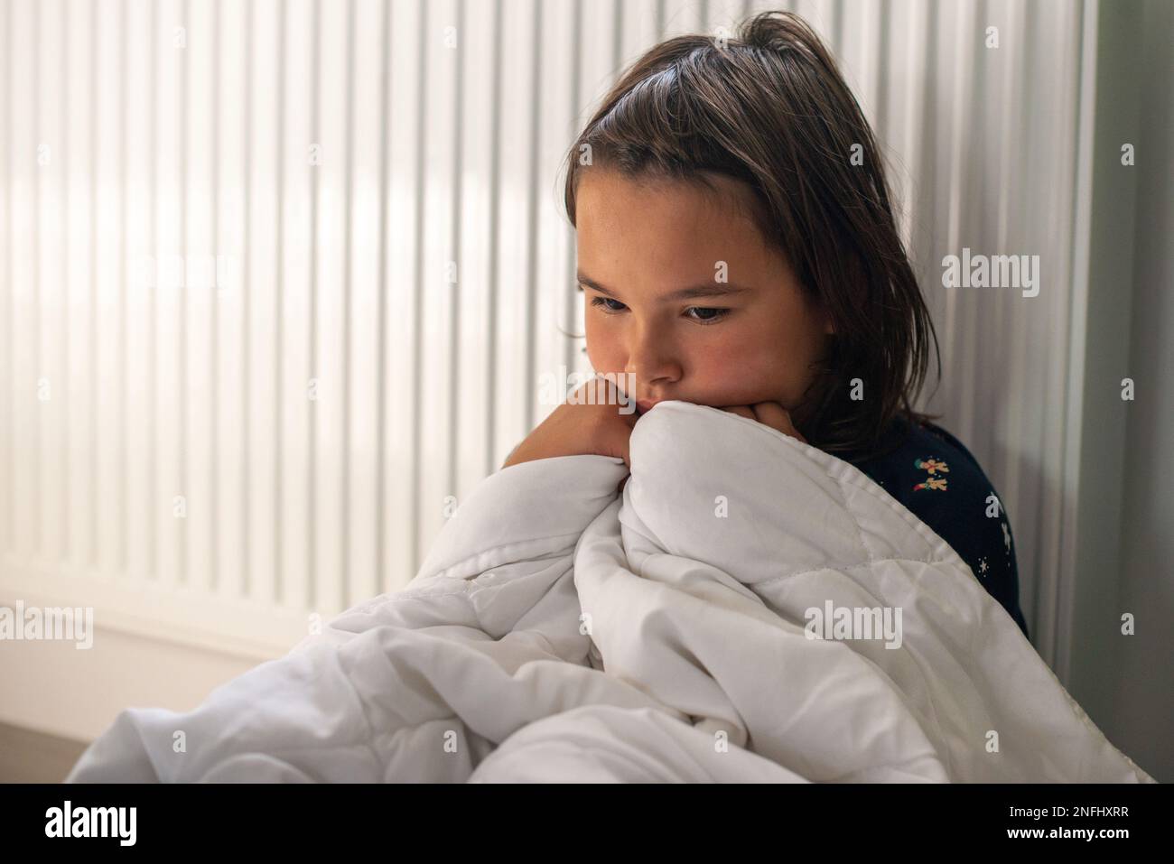 Young girl sitting with a blanket on her next to a radiator to warm ...