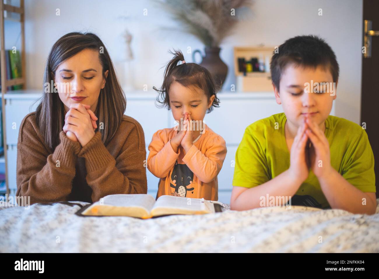 mother and her little girl and boy praying at the edge of the bed in ...