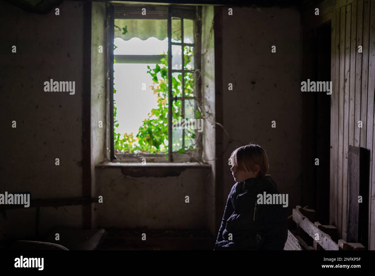 A young boy inside a room in an old crumbling house with an open window ...