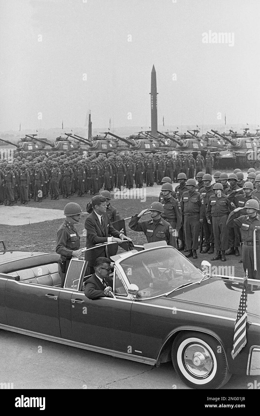President John F. Kennedy turns to look at armored vehicles as he rides ...
