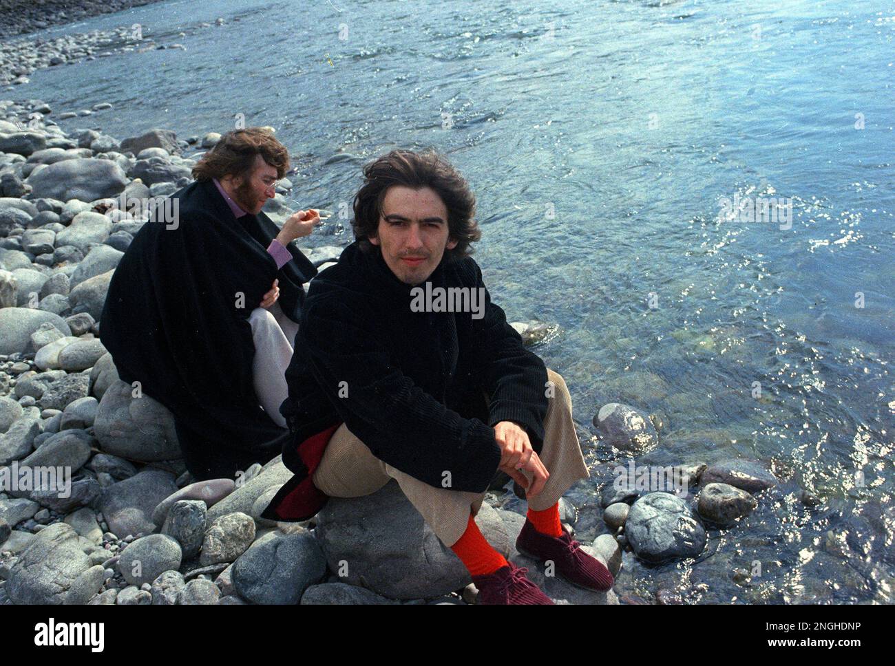 Beatles George Harrison and John Lennon, background, sit on rocks by a ...