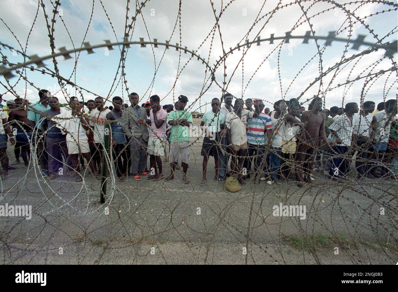 Scores of Haitian refugee men stand behind a razor wire enclosed camp ...
