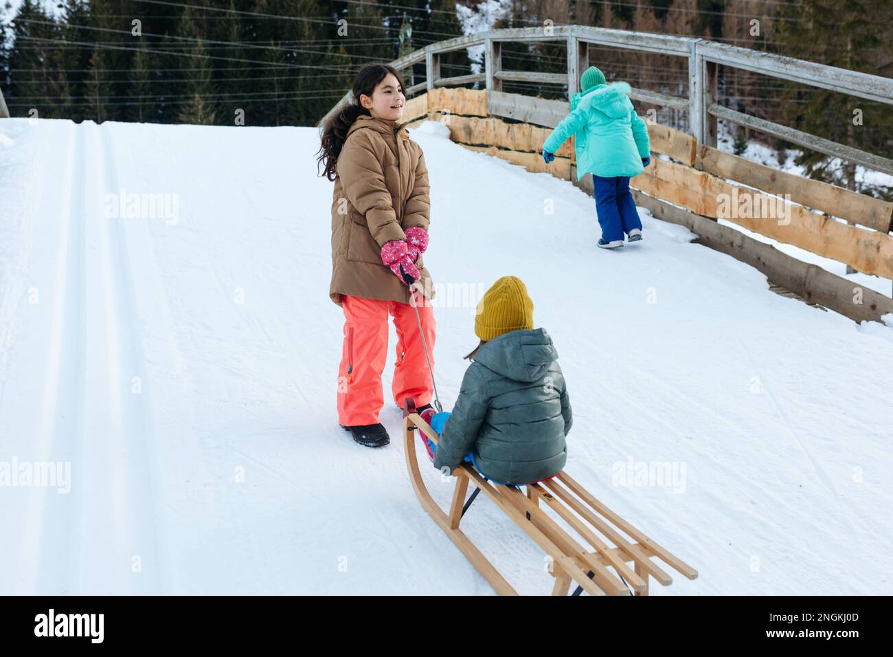 three kids going up to snow slope with small sister on wooden sled ...