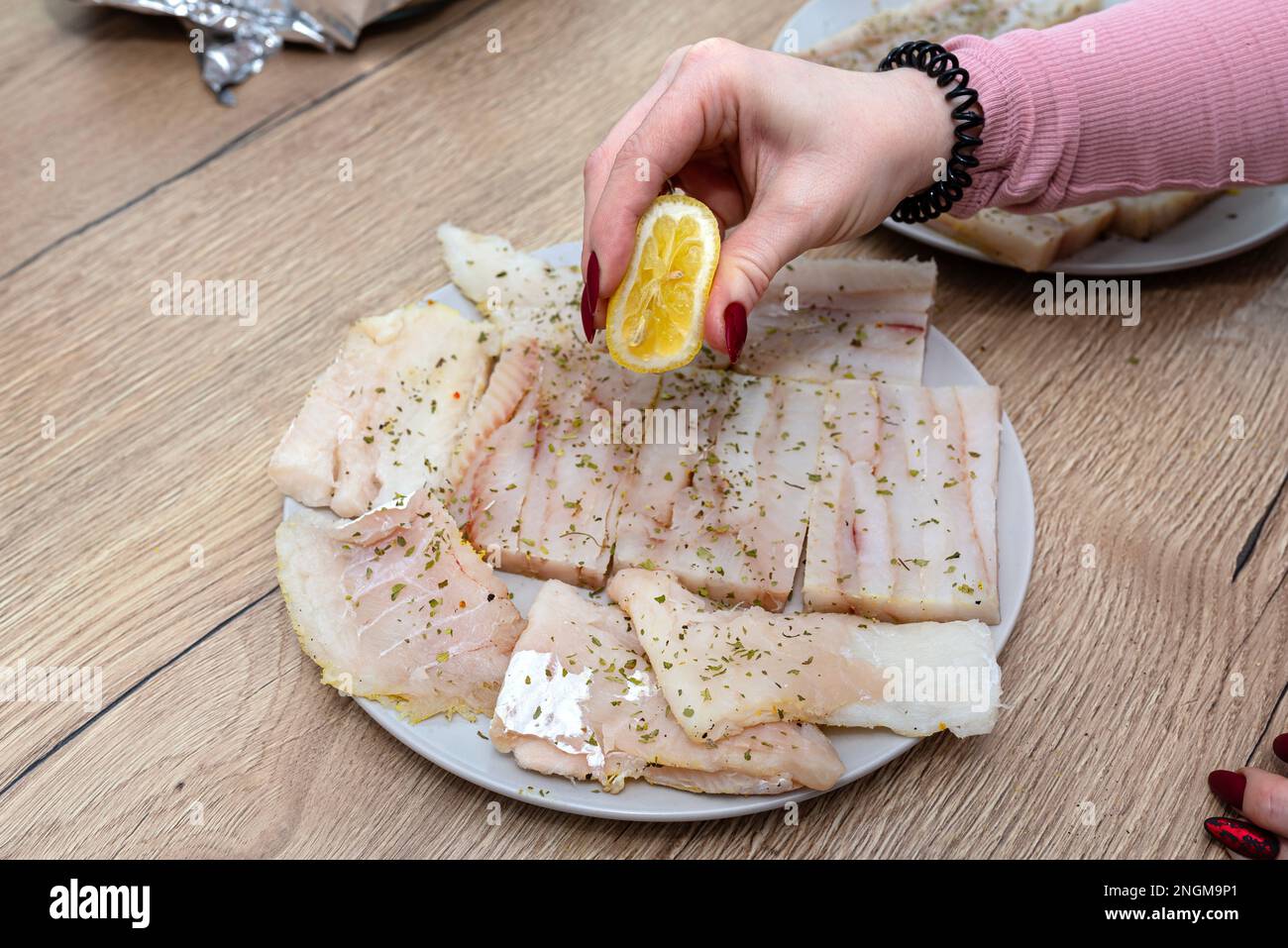 A woman squeezes lemon on a fresh cod fillet cut into squares ...