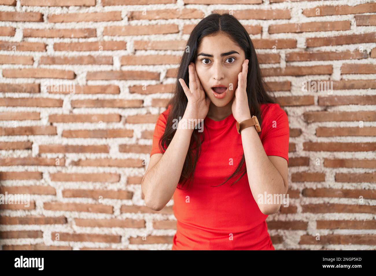 Young teenager girl standing over bricks wall afraid and shocked, surprise and amazed expression ...