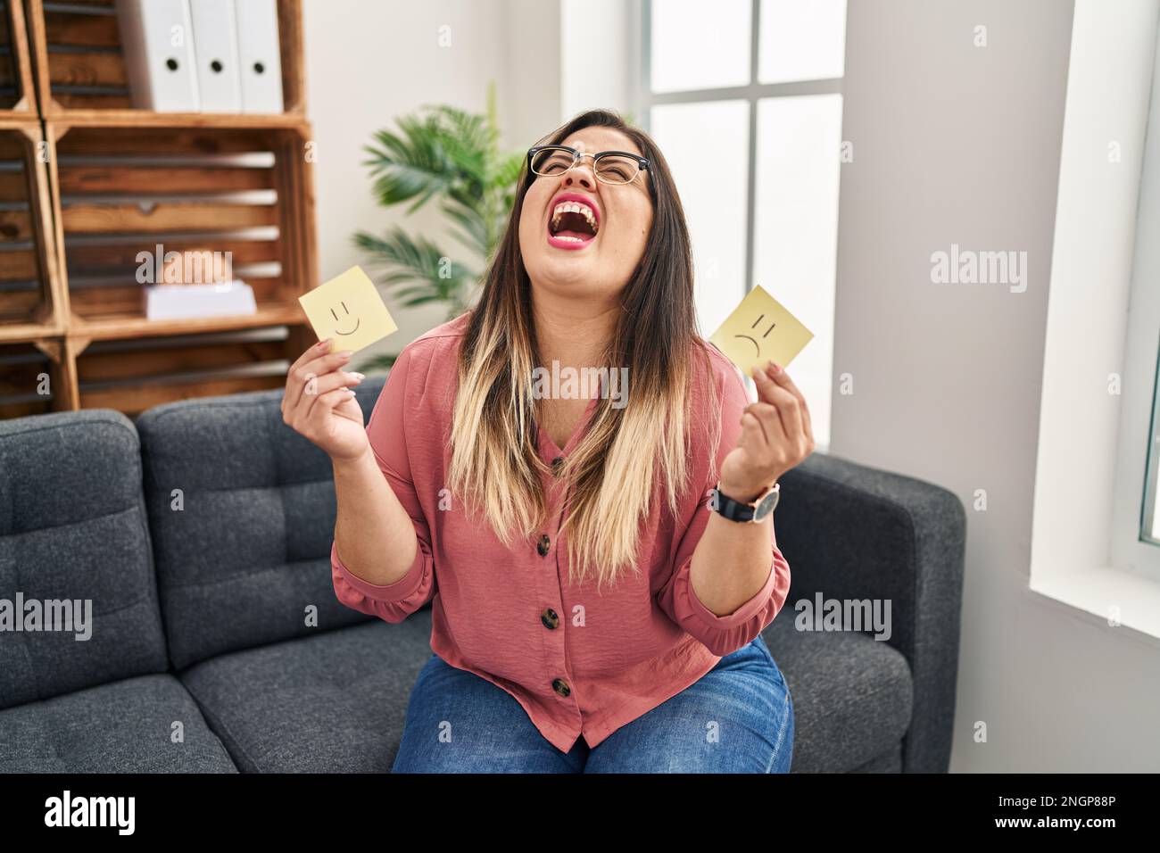Young hispanic woman holding sad to happy emotion paper angry and mad screaming frustrated and ...
