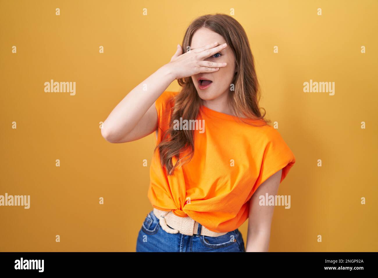 Caucasian woman standing over yellow background peeking in shock ...