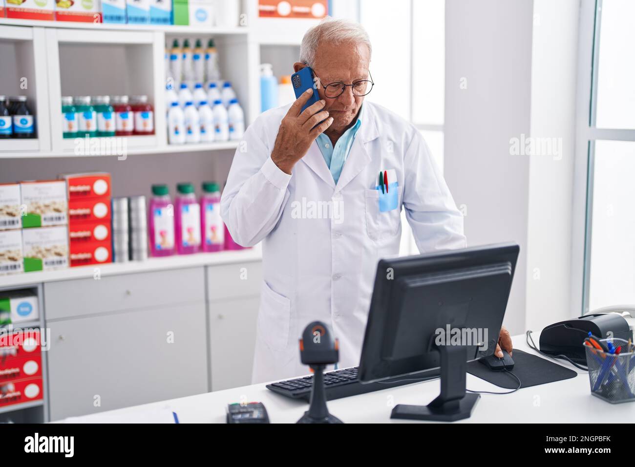 Senior grey-haired man pharmacist talking on smartphone using computer ...