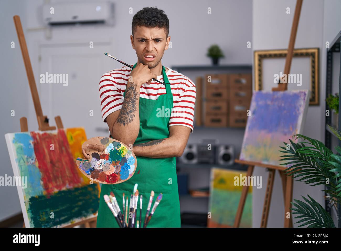Brazilian young man holding painter palette at artist studio clueless ...