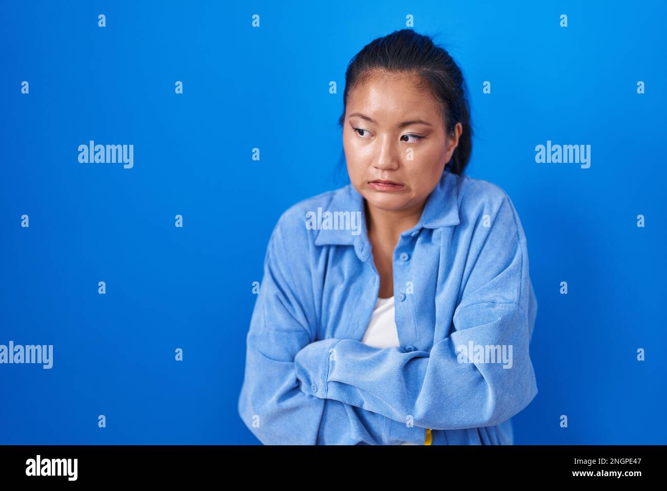 Asian young woman standing over blue background shaking and freezing ...