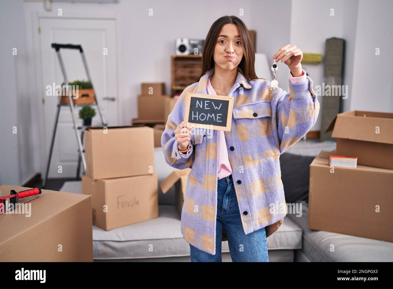 Young brunette woman holding keys and blackboard with new home text puffing cheeks with funny ...