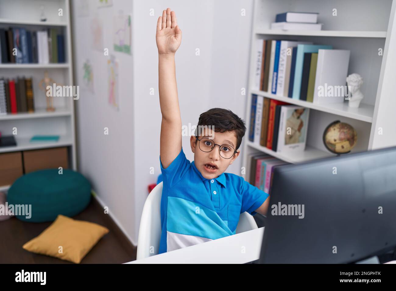 Adorable hispanic boy student using computer with hand raised up at ...