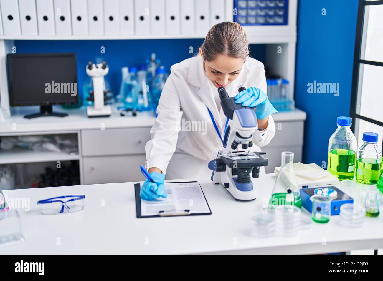 Young woman scientist using microscope writing on report at laboratory ...