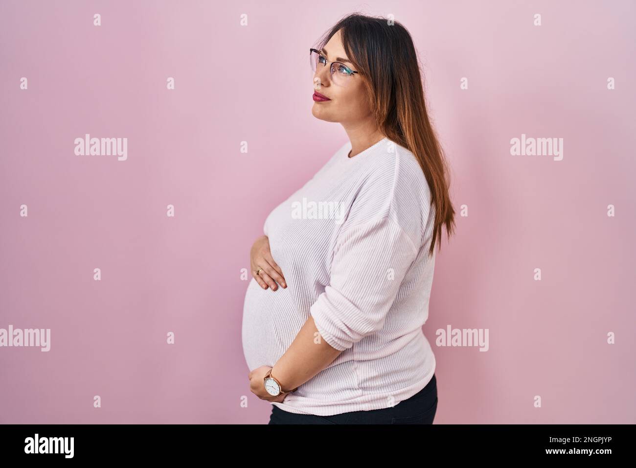 Pregnant woman standing over pink background looking to side, relax ...