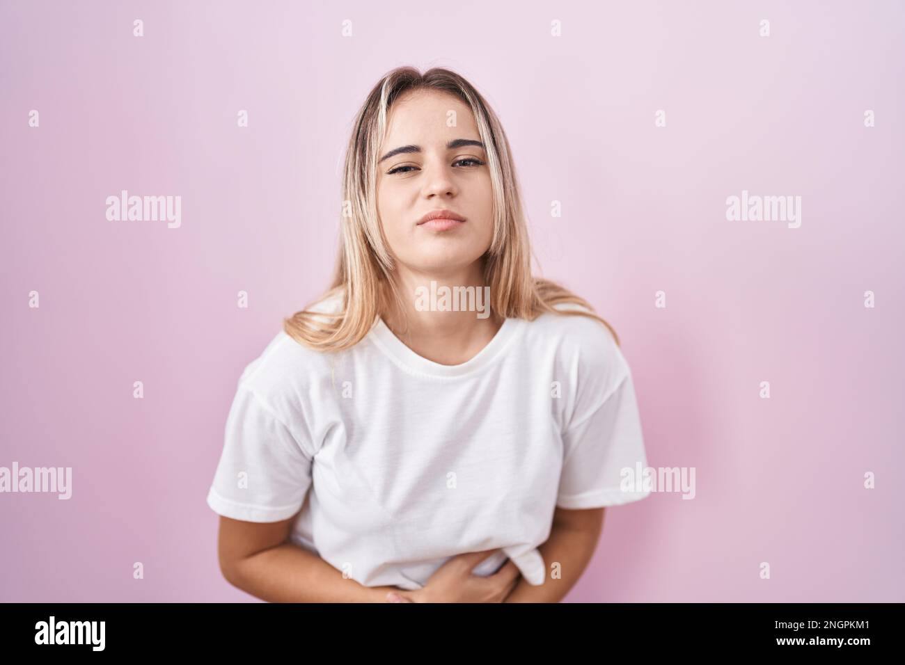 Young blonde woman standing over pink background with hand on stomach ...