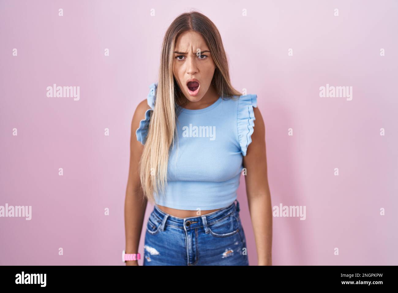 Young blonde woman standing over pink background in shock face, looking ...
