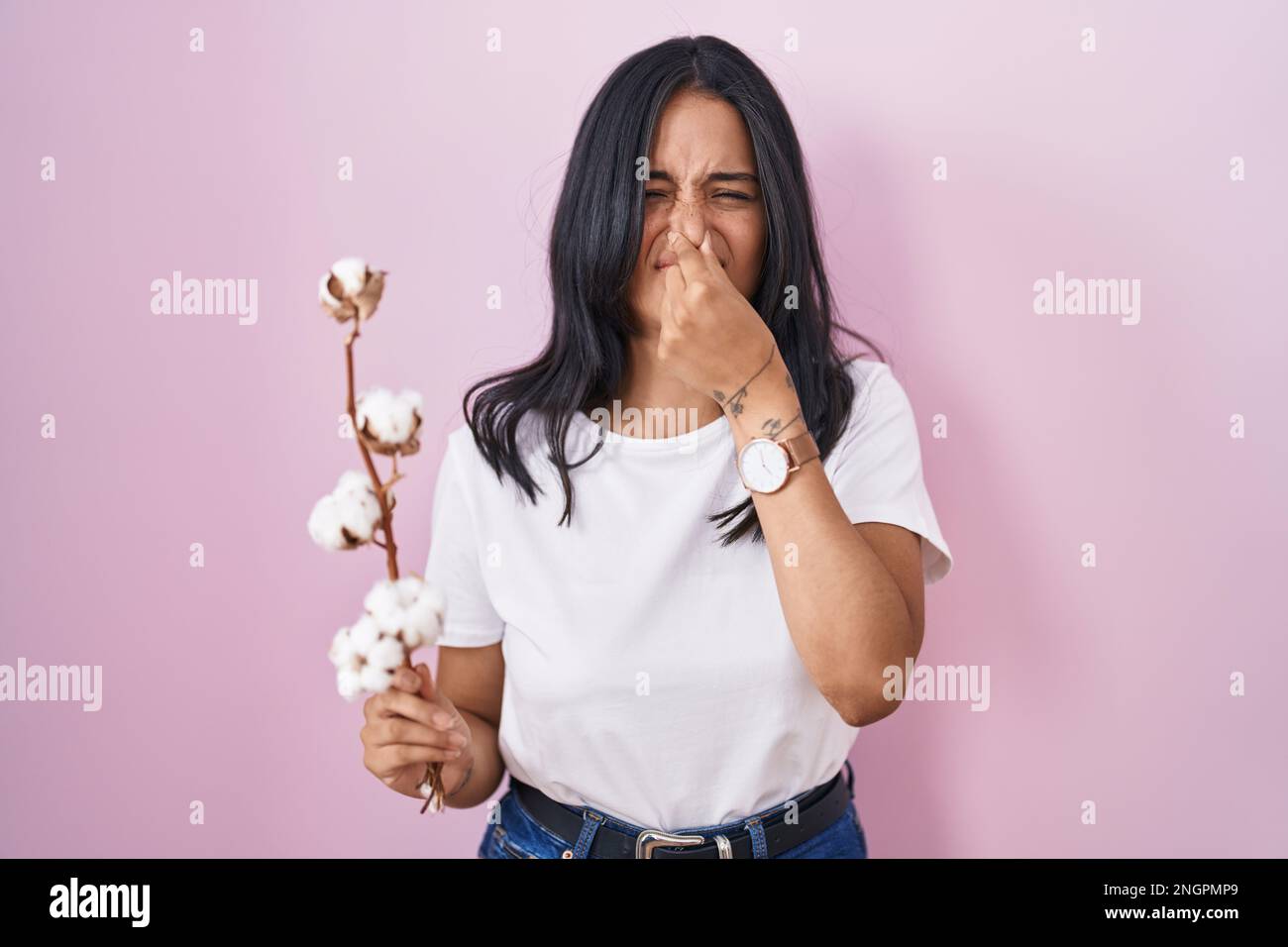 Brunette woman standing over pink background smelling something stinky ...