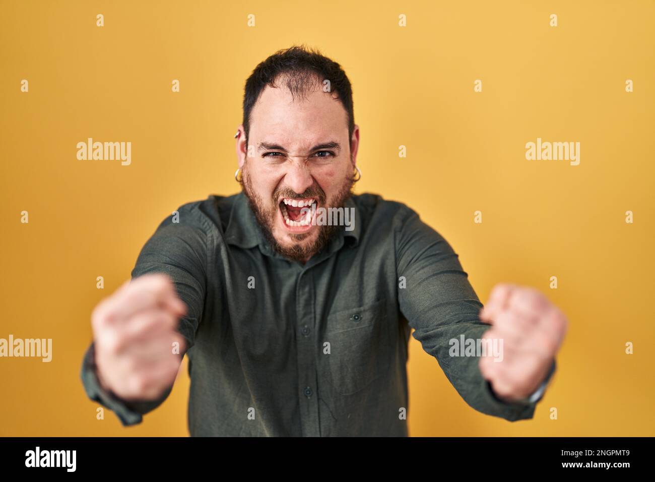Plus size hispanic man with beard standing over yellow background angry ...