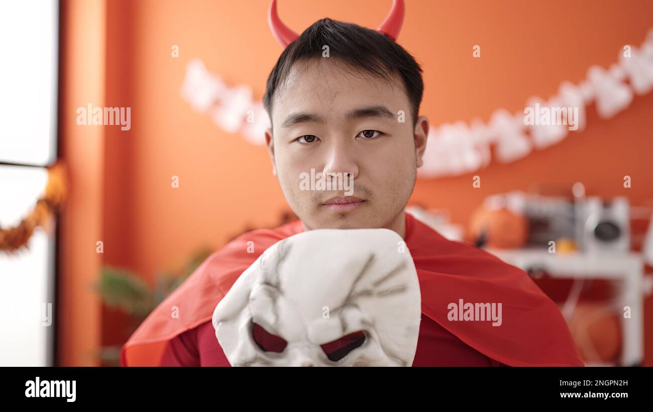 Young chinese man wearing devil costume holding skull mask at home ...