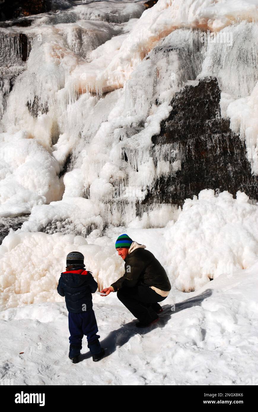 A father and son walk on an ice sheet to view a flowing waterfall ...