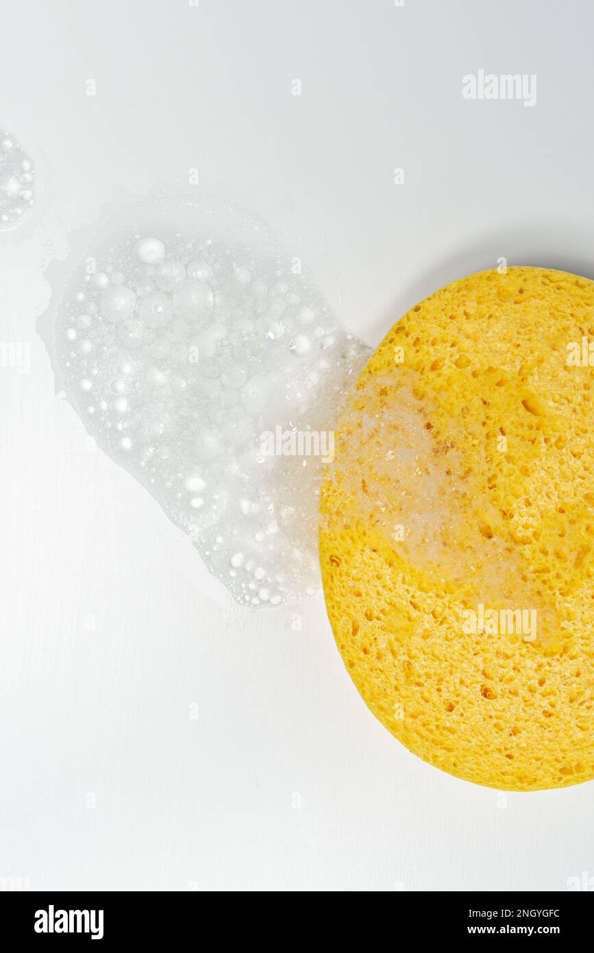 A yellow synthetic bath sponge with soap foam on a white surface seen ...
