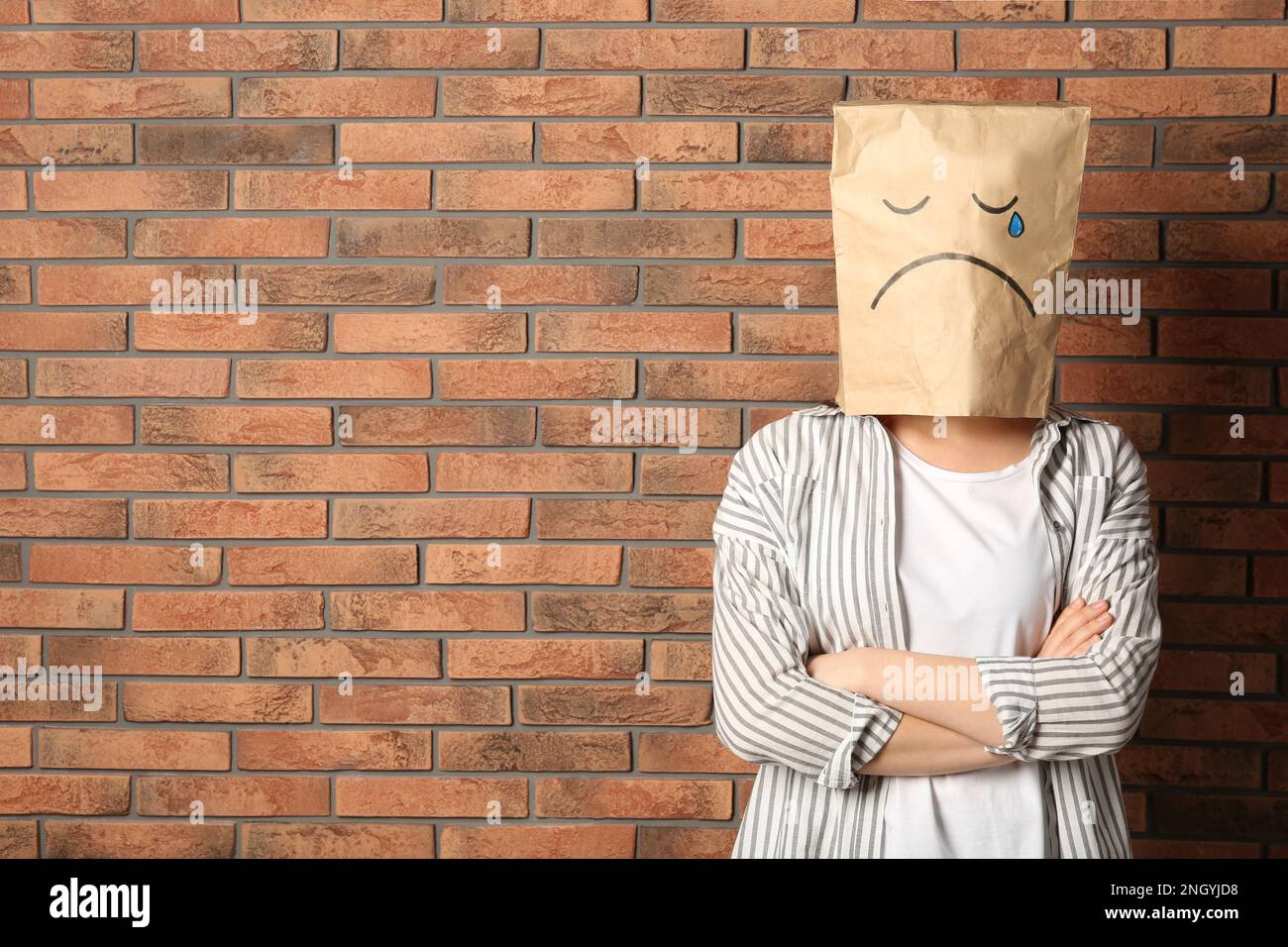 Woman wearing paper bag with drawn sad face near brick wall. Space for ...