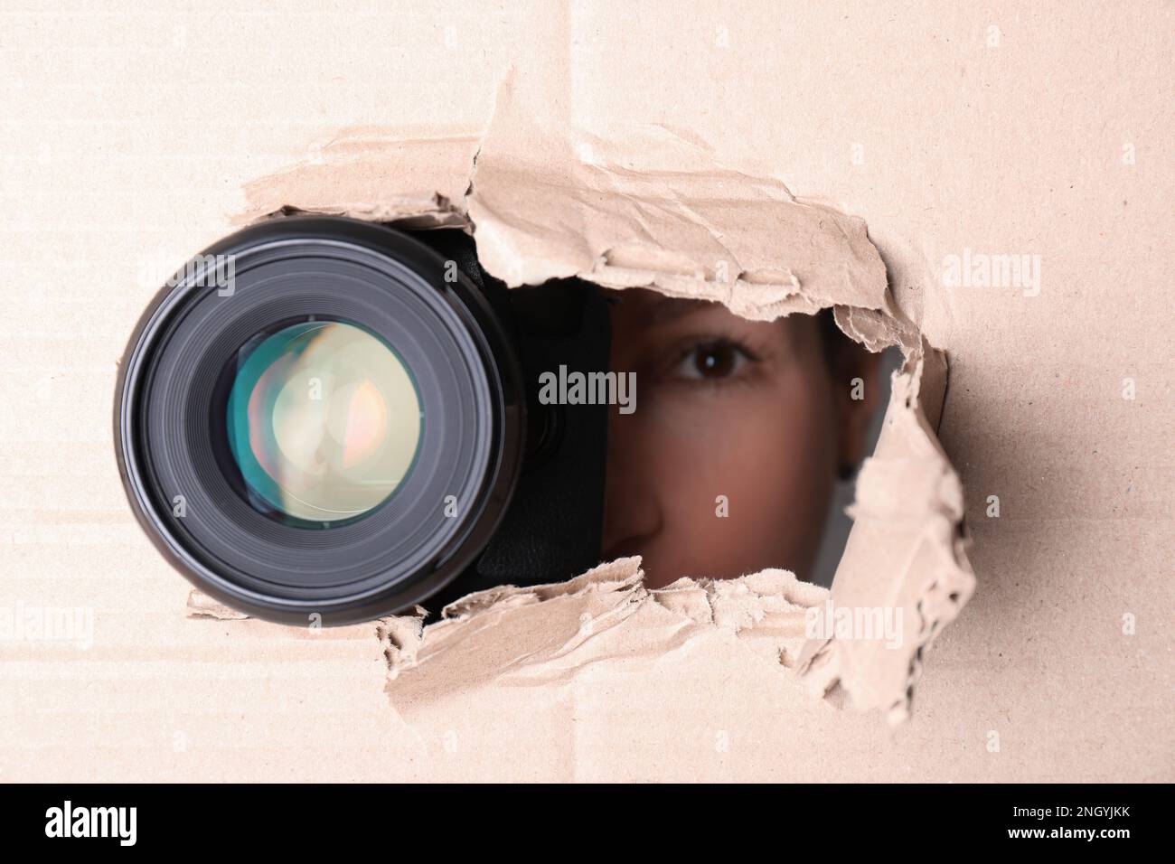 Hidden woman with camera spying through torn hole in cardboard, closeup ...