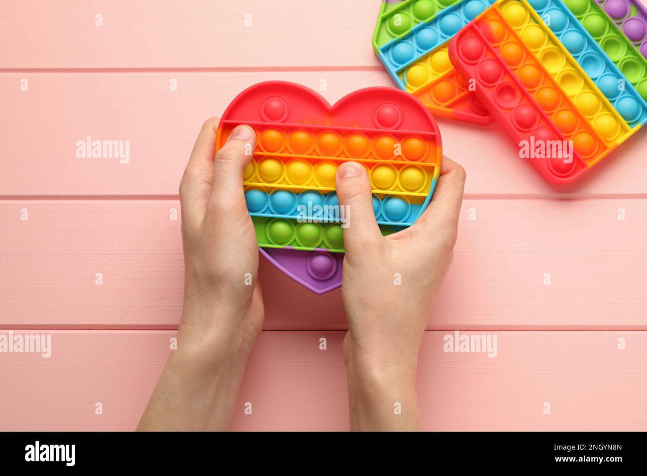 Woman using heart shaped pop it fidget toy at pink wooden table, top ...